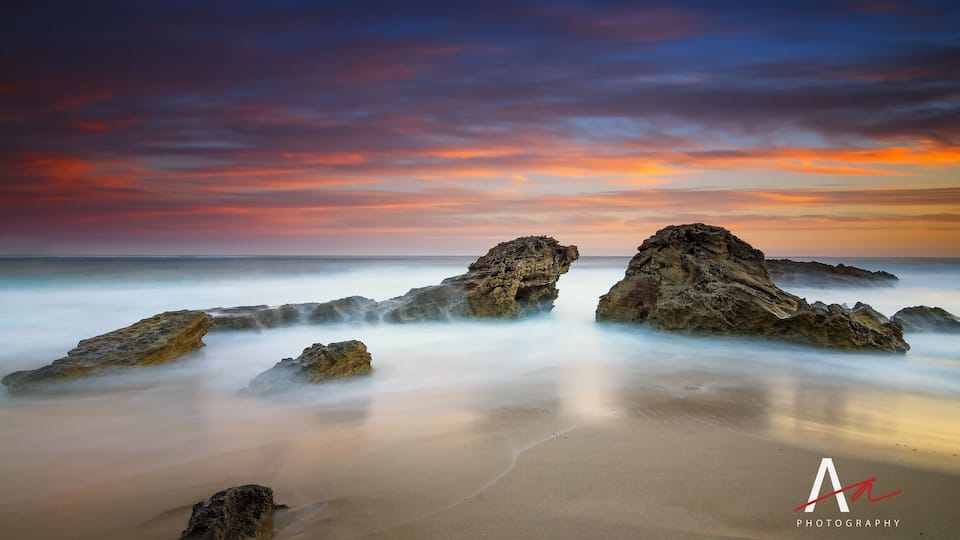 Uma praia com águas calmas e formações rochosas sob um céu vibrante ao pôr do sol.