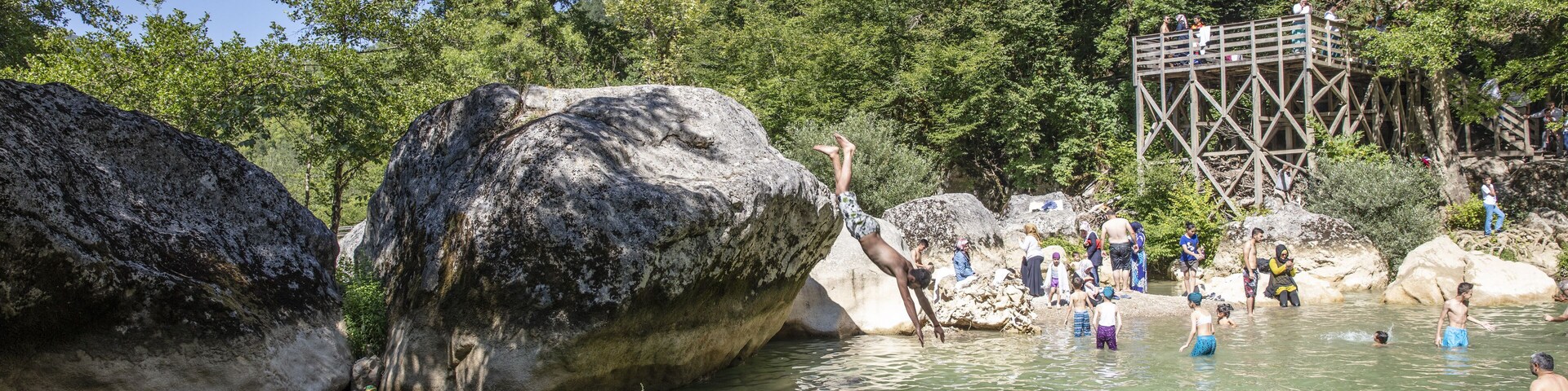 Uma piscina natural rodeada por pedras grandes e árvores, com pessoas a nadar e uma plataforma de madeira para observação.