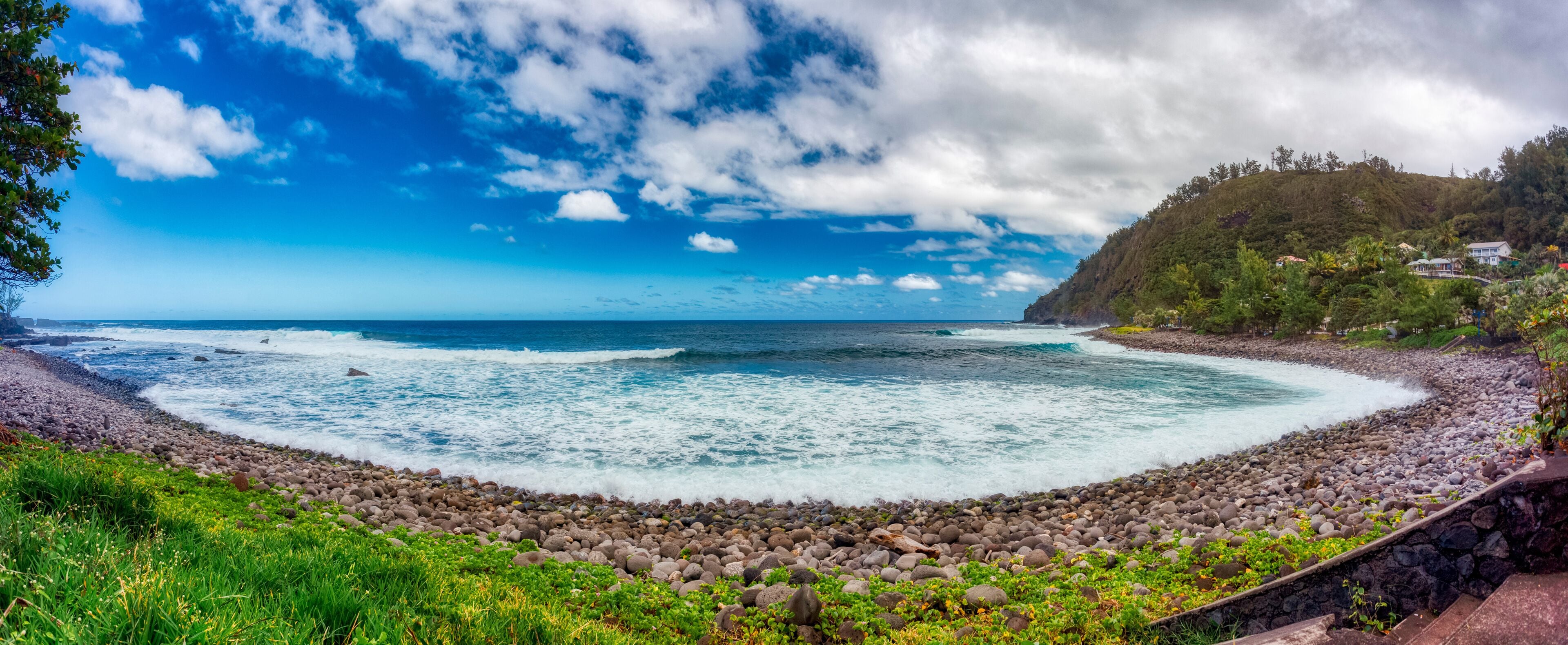 Uma praia rochosa com ondas quebrando na costa, um céu azul límpido e um morro com vegetação e edifícios ao fundo.