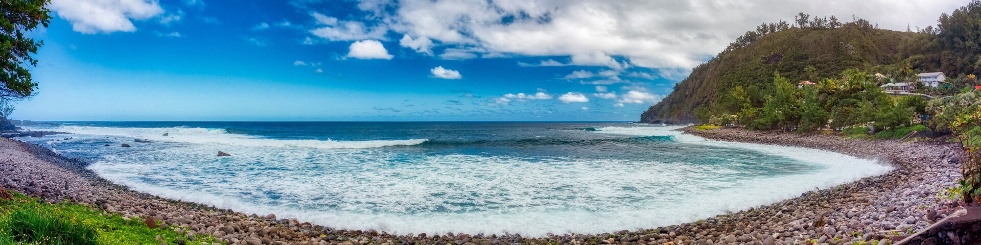 Uma praia rochosa com ondas quebrando na costa, um céu azul límpido e um morro com vegetação e edifícios ao fundo.