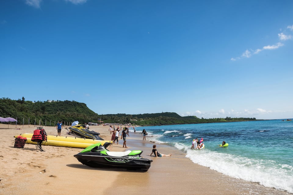 Uma praia com jet skis, pessoas na água e um céu azul límpido.