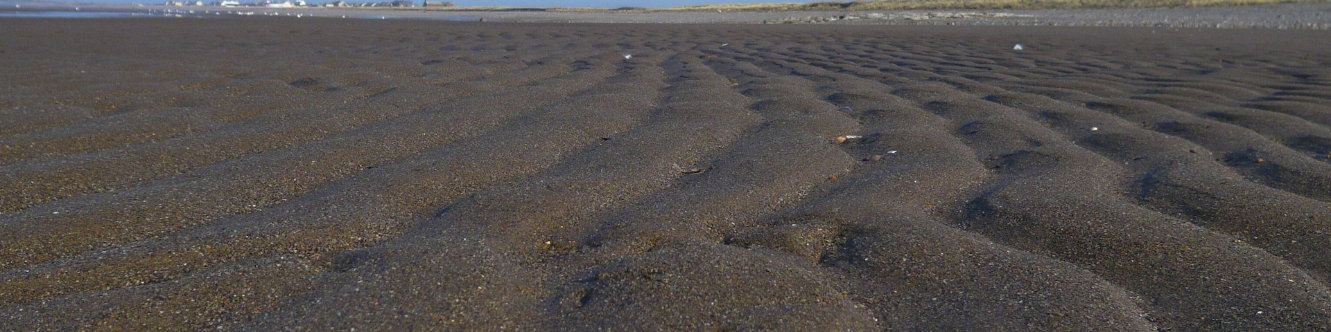 Uma praia arenosa com ondulações na areia e edifícios ao longe.
