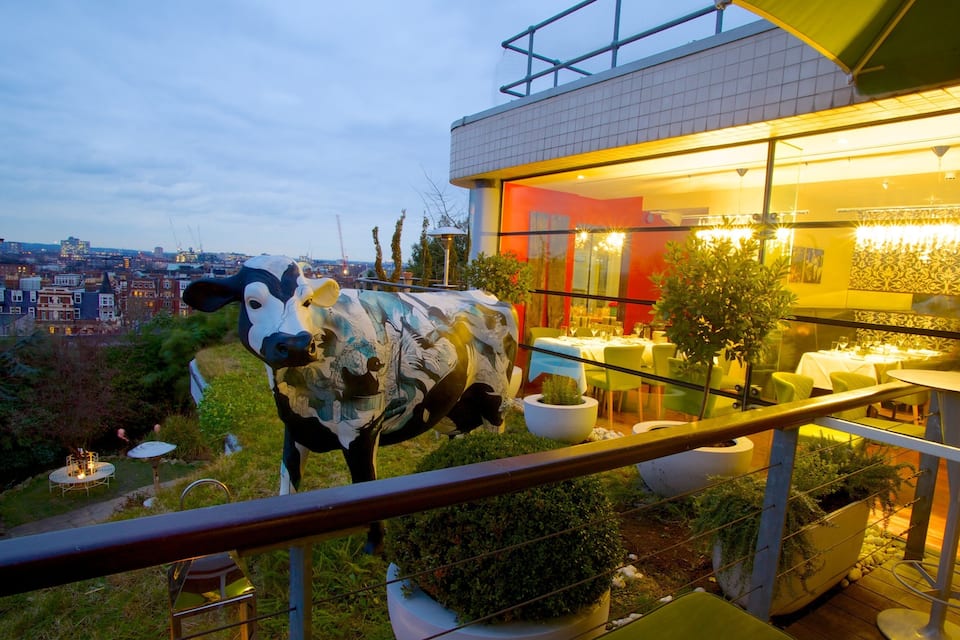 Paisagem urbana com uma grande escultura de vaca colorida em um jardim, com vista para um edifício moderno com área de estar ao ar livre.