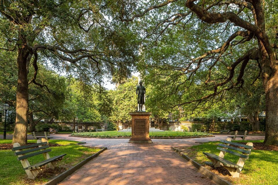 Um parque com uma estátua, bancos e árvores.