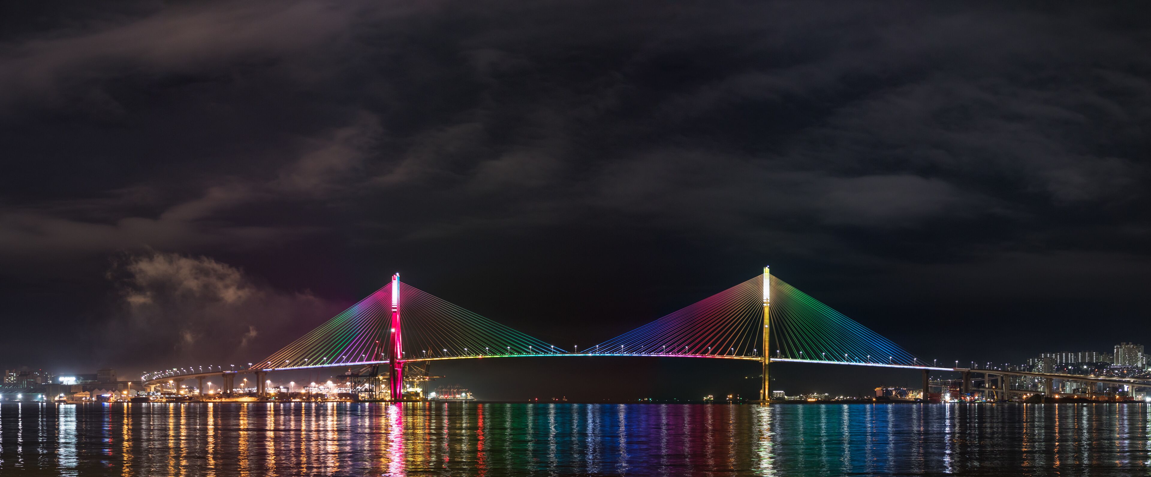 Vista noturna de uma ponte com luzes coloridas refletindo na água.