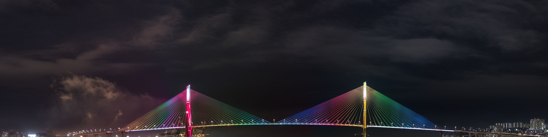 Vista noturna de uma ponte com luzes coloridas refletindo na água.