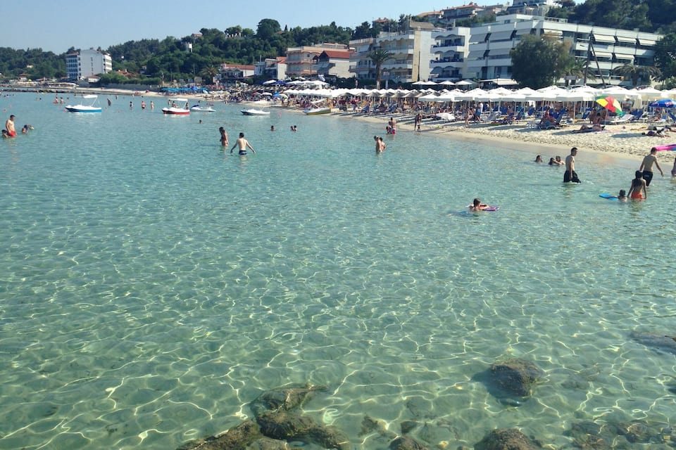 Uma praia movimentada com águas claras e rasas, e áreas rochosas.