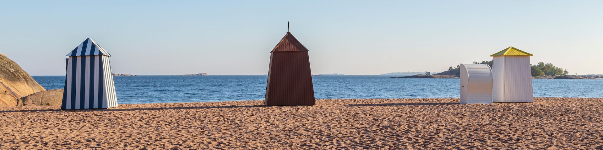 Uma praia arenosa com três construções distintas: um quiosque listrado, um abrigo cônico marrom e um casebre branco com telhado amarelo.
