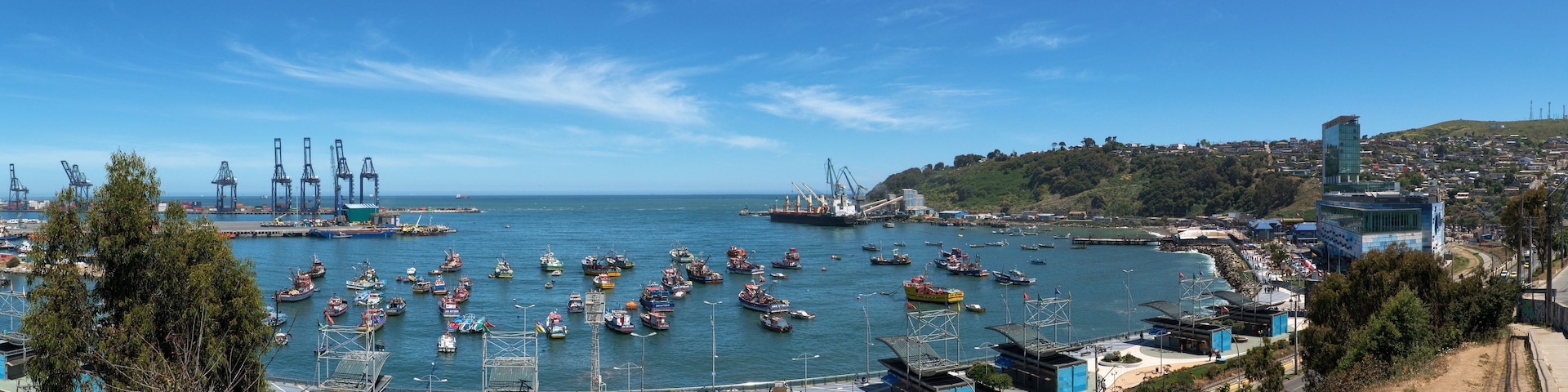 Um porto com muitos barcos, guindastes e uma paisagem urbana ao fundo.