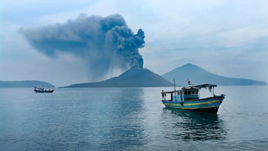 Dois barcos em águas calmas com um vulcão ao fundo, expelindo fumaça.