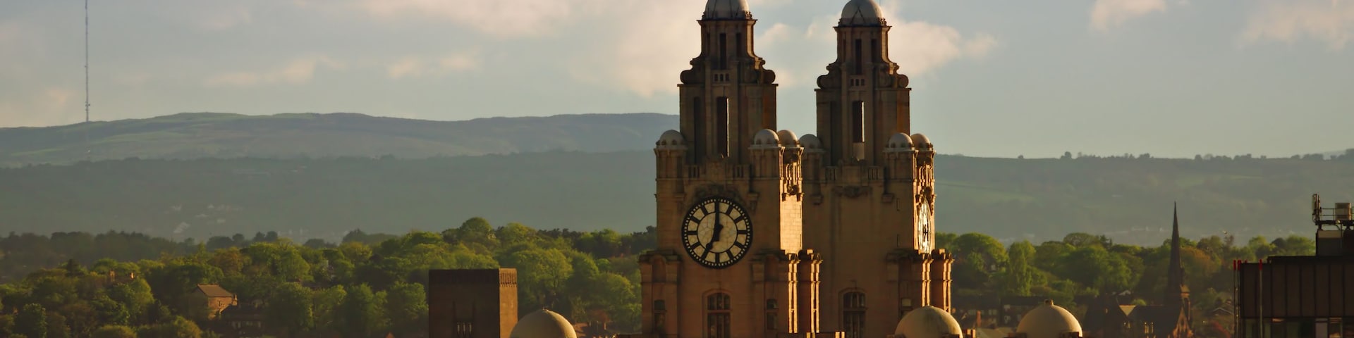 Uma catedral histórica com duas torres gêmeas e um grande relógio, com o cenário de um horizonte urbano e colinas ao longe.