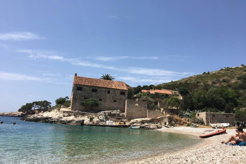 Uma paisagem costeira com um edifício de pedra, águas cristalinas e uma praia de pedras.