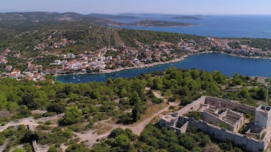 Vista aérea de uma vila costeira com uma fortaleza histórica, vegetação densa e mar azul cristalino.