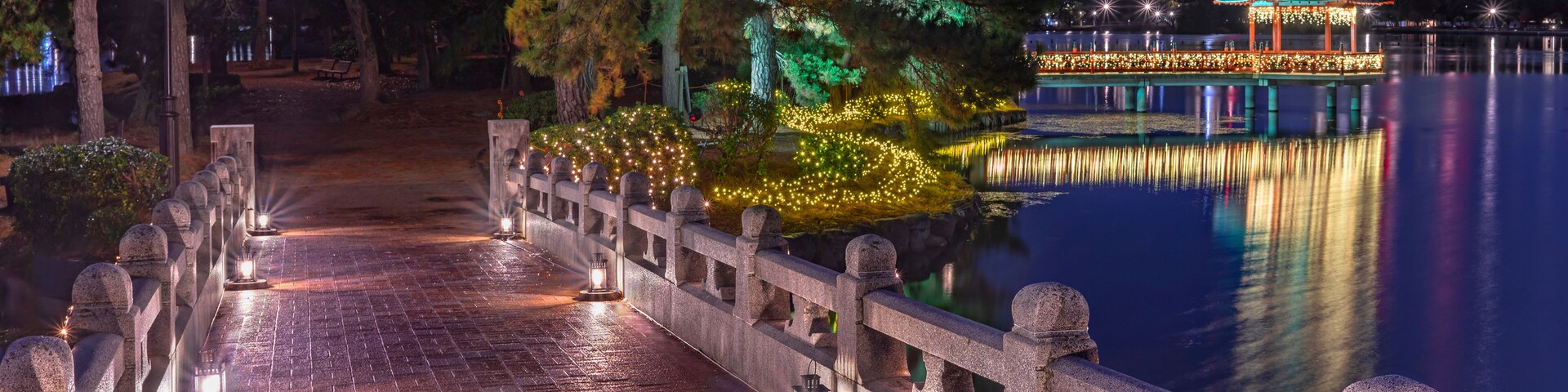 Uma ponte de pedra com lanternas, um coreto iluminado com luz verde e o horizonte urbano ao fundo.