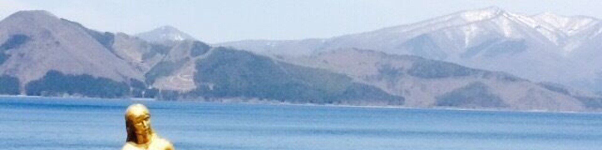Uma estátua dourada sobre uma rocha, perto de um lago, com montanhas ao fundo.