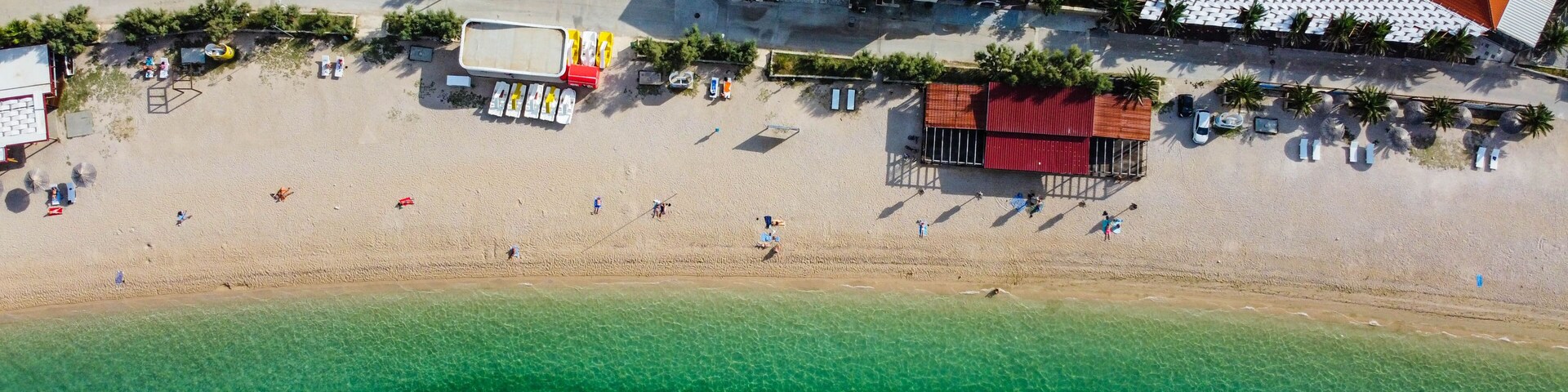 Vista aérea de uma praia com águas turquesa cristalinas, uma fileira de guarda-sóis de praia e um edifício com telhado vermelho.