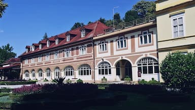 Um edifício histórico com janelas em arco e telhado vermelho, rodeado por jardins bem cuidados e um céu azul límpido.