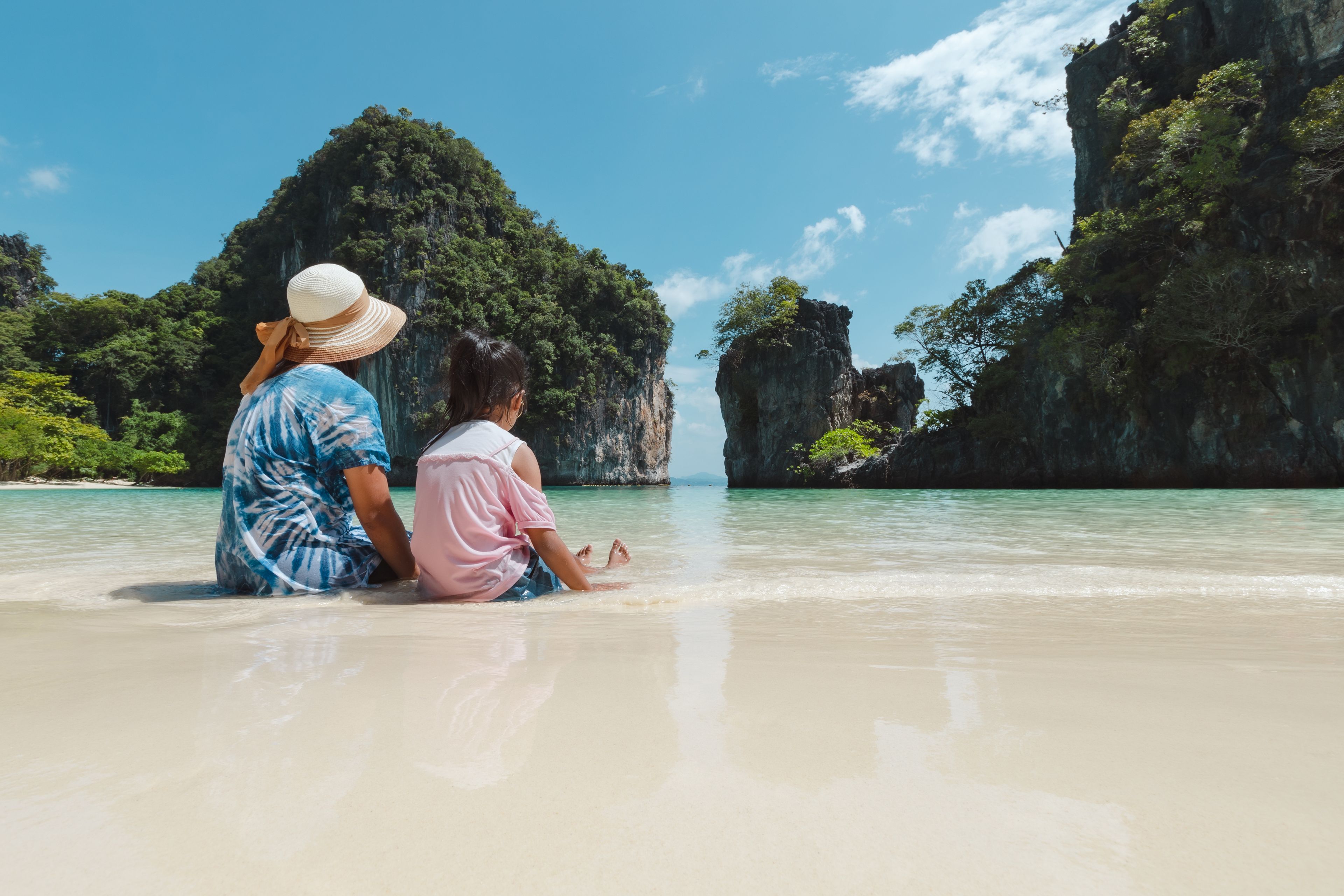 Duas pessoas sentadas numa praia com águas turquesa translúcidas e grandes formações rochosas ao fundo.