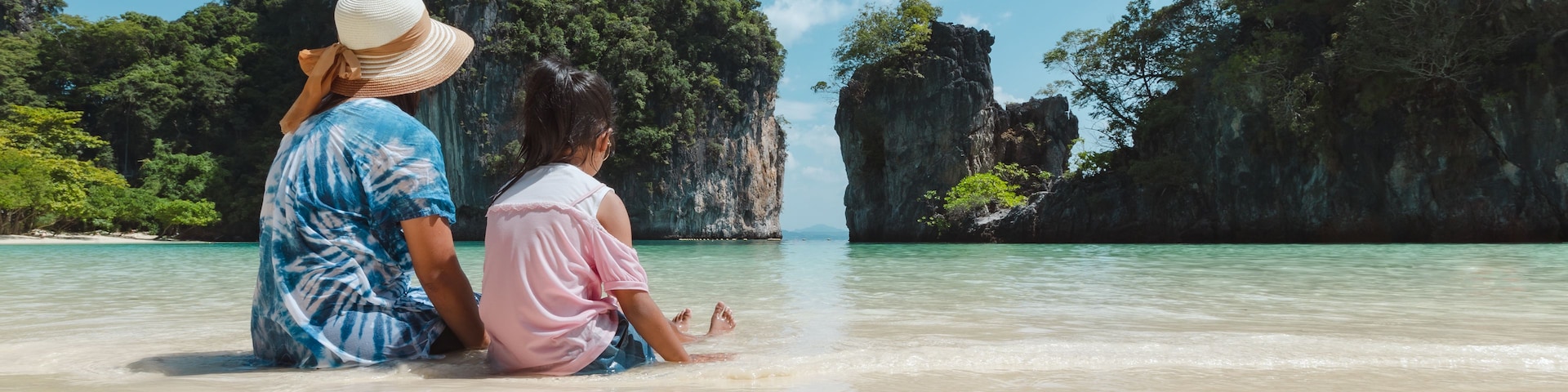 Duas pessoas sentadas numa praia com águas turquesa translúcidas e grandes formações rochosas ao fundo.