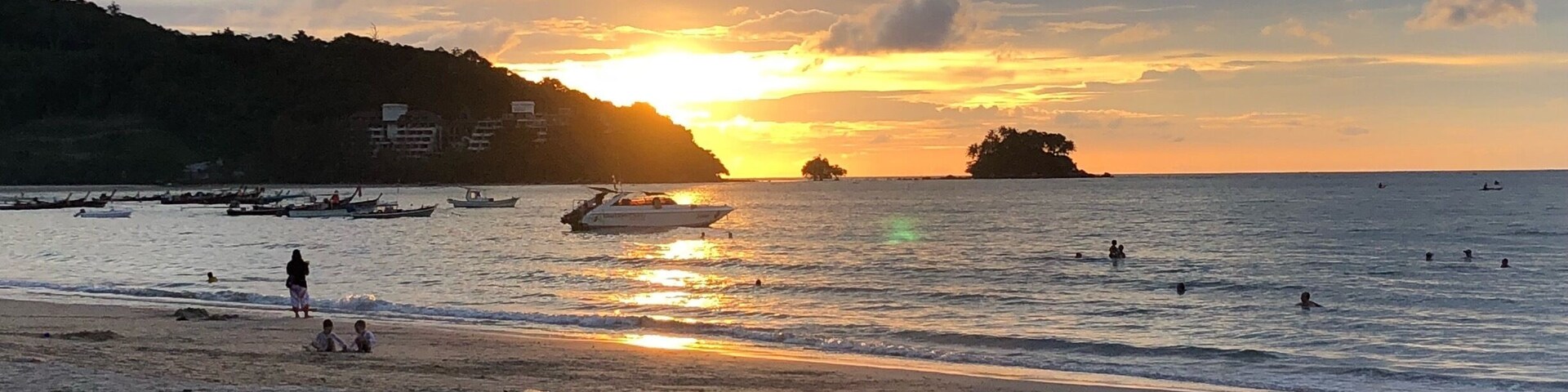 Praia ao pôr do sol com barcos, pessoas nadando e uma colina arborizada ao fundo.