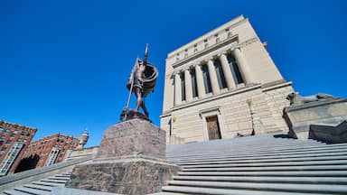 Uma estátua sobre um pedestal em frente a um edifício com colunas e uma torre do relógio.