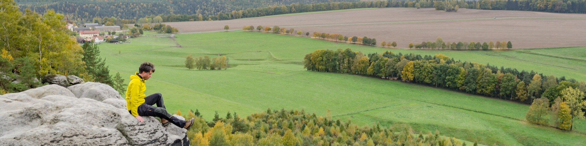 Uma pessoa com uma jaqueta amarela sentada em uma rocha, com vista para um vale com campos e colinas.