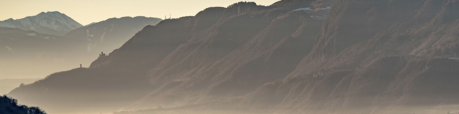 Paisagem montanhosa e neblinosa com uma cidade aninhada na base, destacando uma torre de igreja imponente e edifícios industriais.