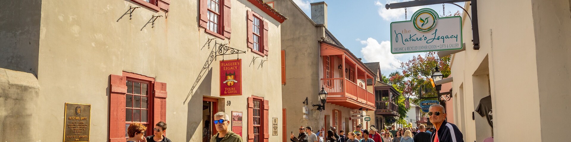 Uma rua movimentada com edifícios históricos, uma placa indicando a loja 'The Nature's Legacy' e uma placa comemorativa para 'Oliver's House'.
