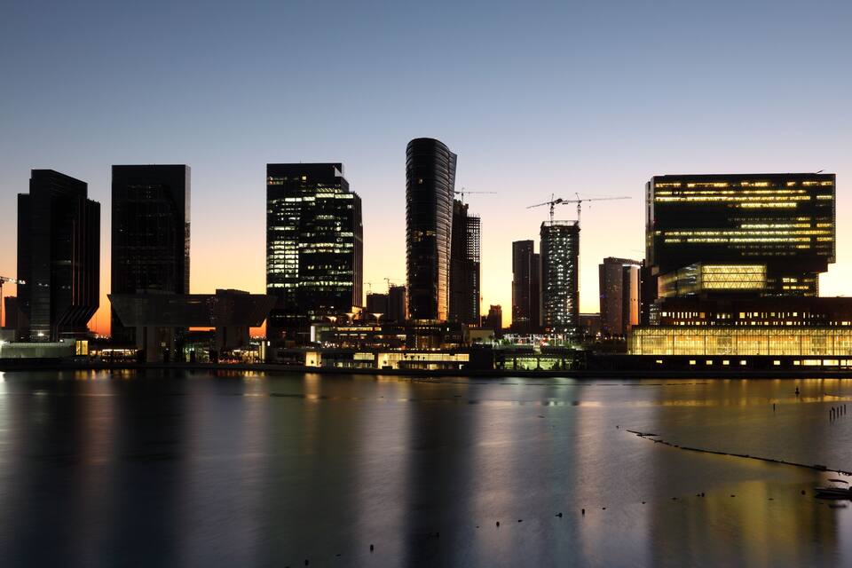 Silhueta urbana ao anoitecer, com edifícios iluminados e um rio calmo refletindo as luzes.