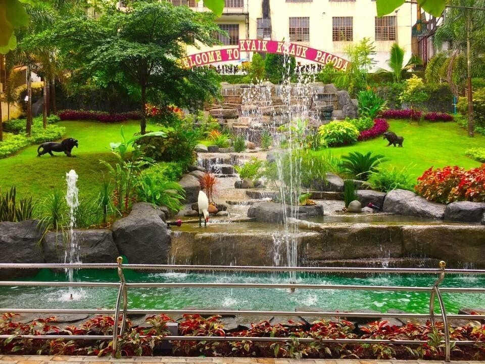 Um jardim paisagístico com uma cascata, uma placa com a mensagem 'Welcome to Kyaik Phauk Pagoda' e estátuas de animais.