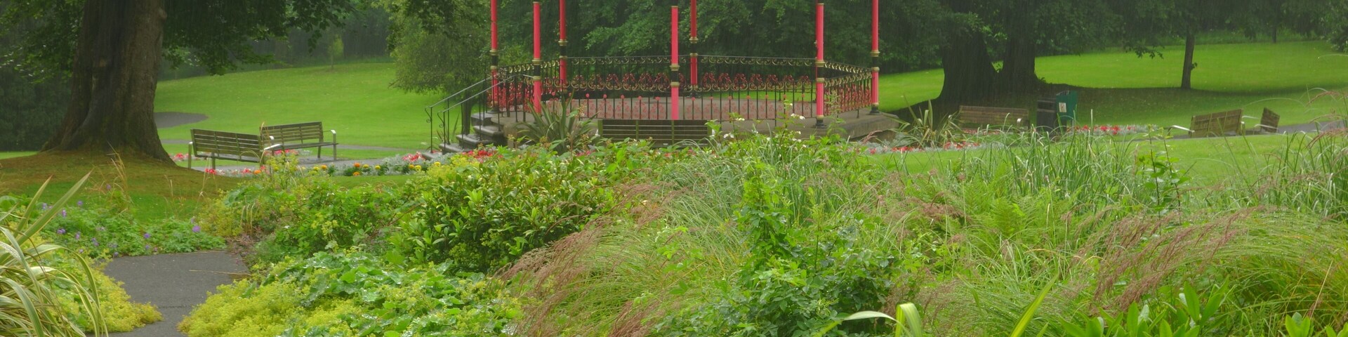 Um gazebo num parque, com um caminho a levá-lo até ele, rodeado por vegetação exuberante e árvores.