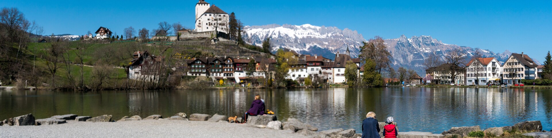 Um lago com casas e um castelo numa colina, rodeado por montanhas.