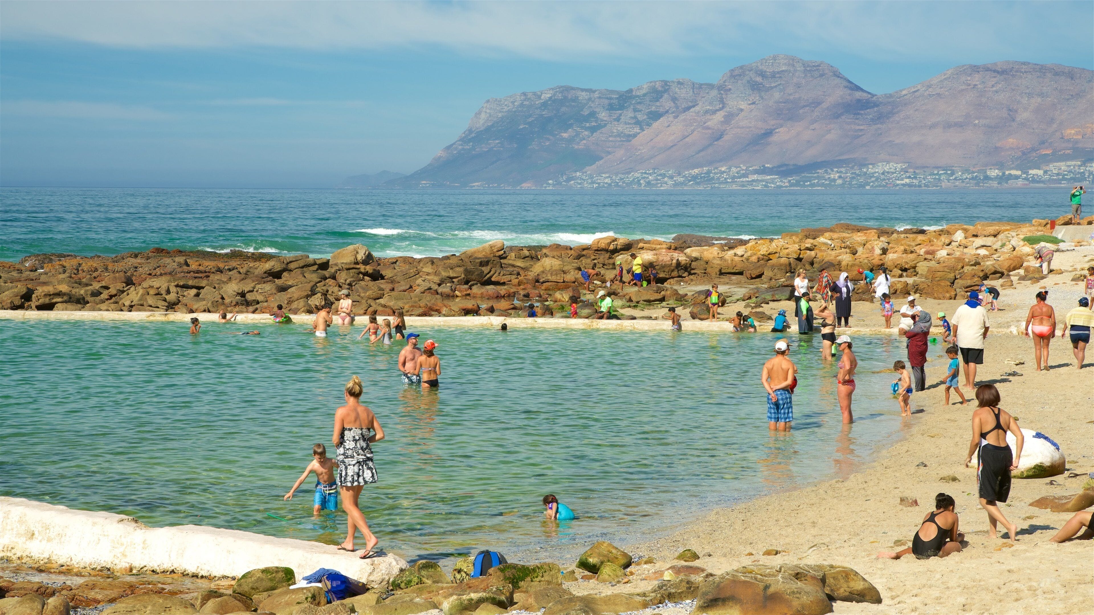 Praia lotada com pessoas nadando e tomando sol, costa rochosa e montanhas ao fundo.