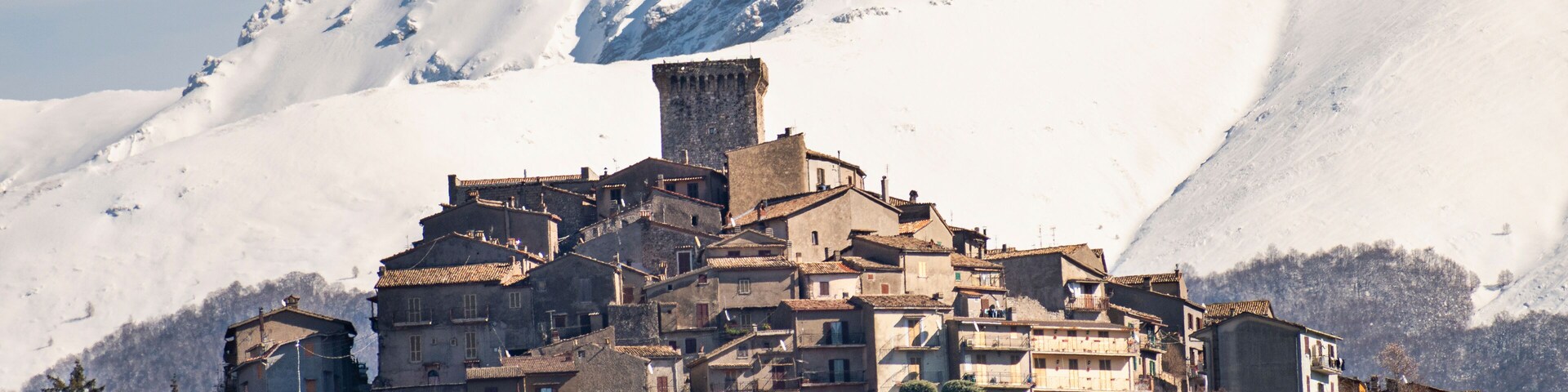 Uma aldeia coberta de neve com edifícios históricos e uma torre imponente, rodeada por montanhas.