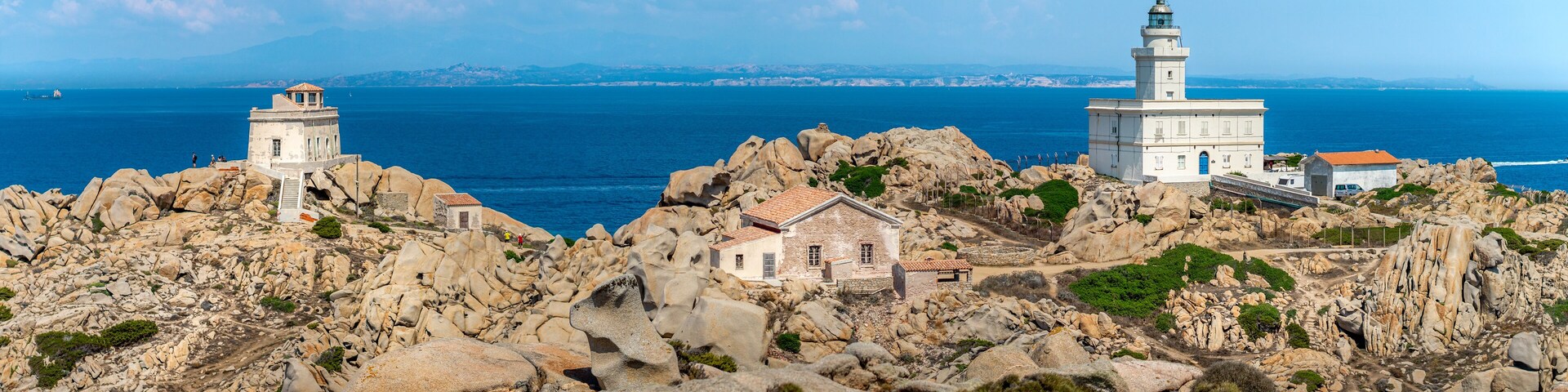 Paisagem costeira com terreno rochoso, um farol e um pequeno edifício.