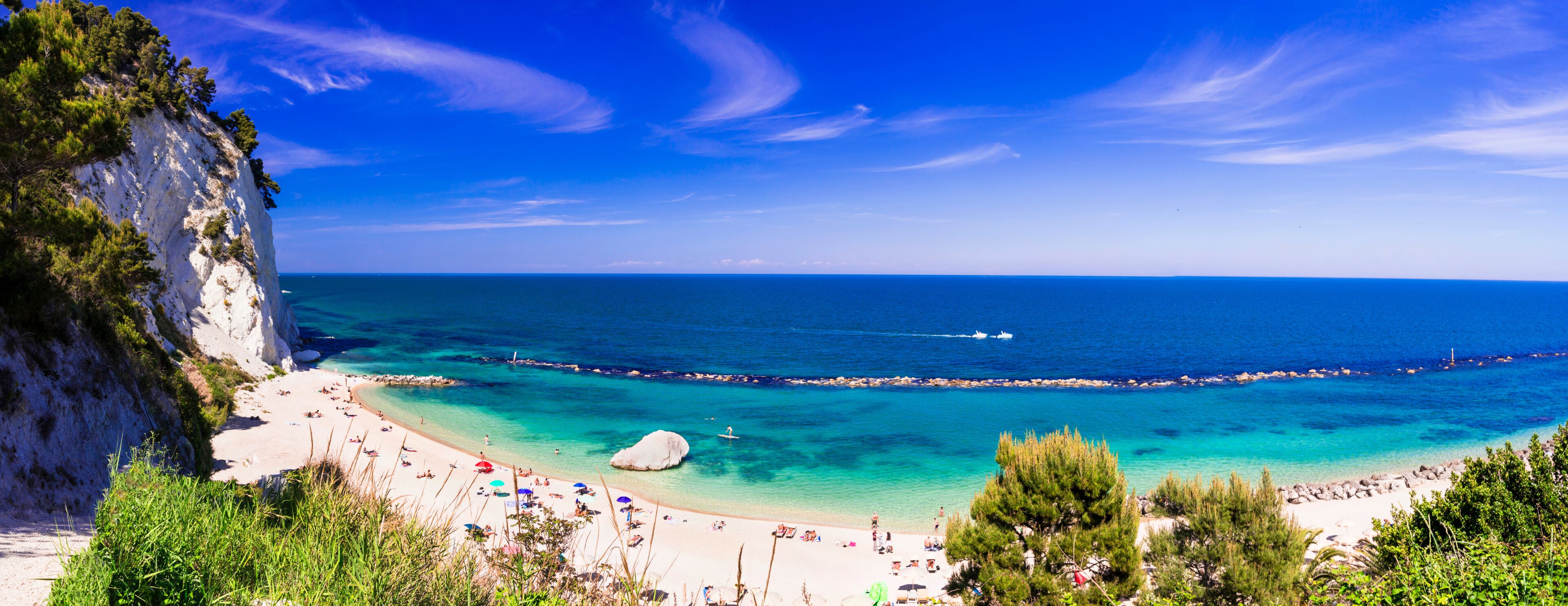 Uma praia costeira com águas turquesa cristalinas, um molhe rochoso e uma faixa de areia branca.