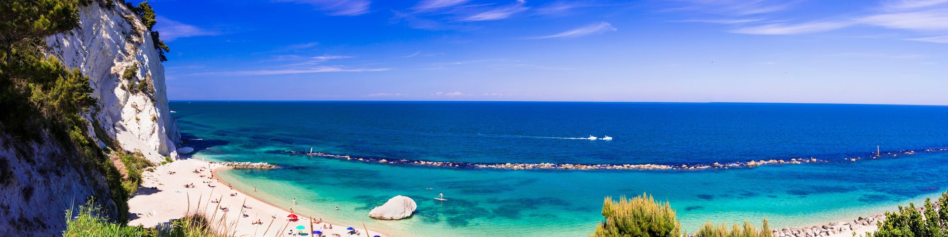 Uma praia costeira com águas turquesa cristalinas, um molhe rochoso e uma faixa de areia branca.