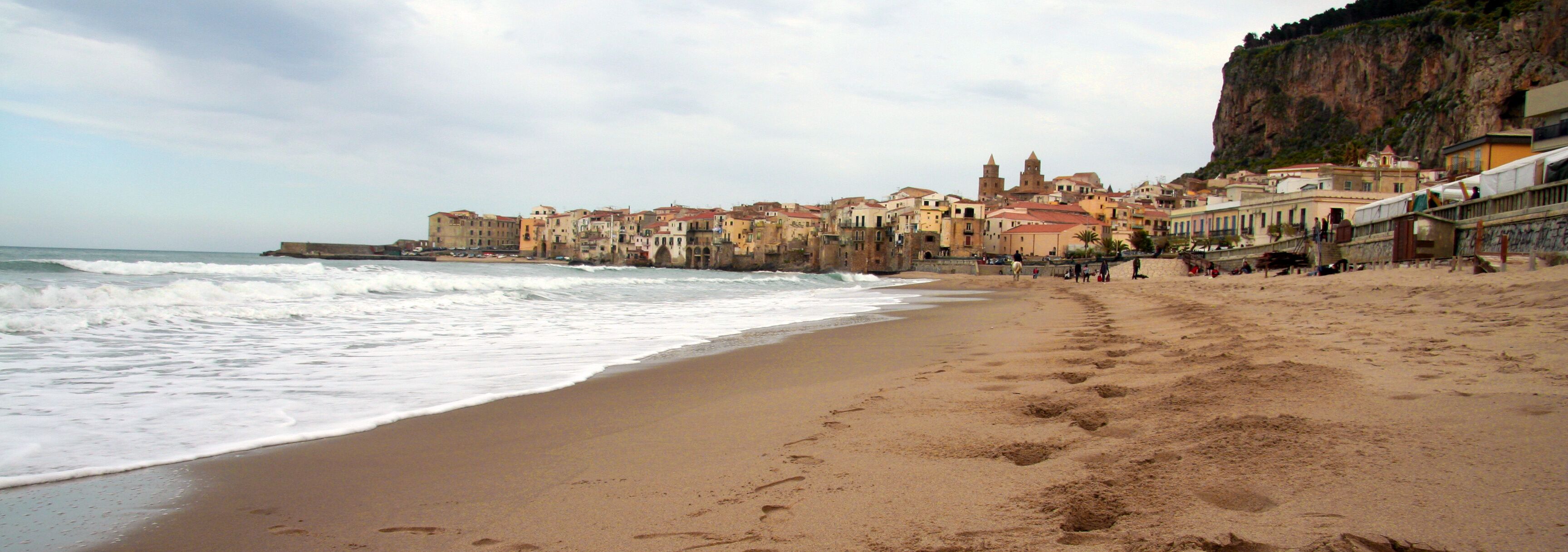 Uma praia arenosa com pegadas, ondas e uma cidade costeira com edifícios históricos.