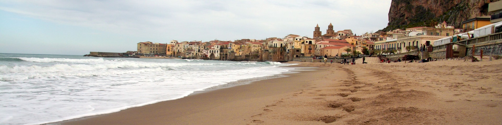 Uma praia arenosa com pegadas, ondas e uma cidade costeira com edifícios históricos.