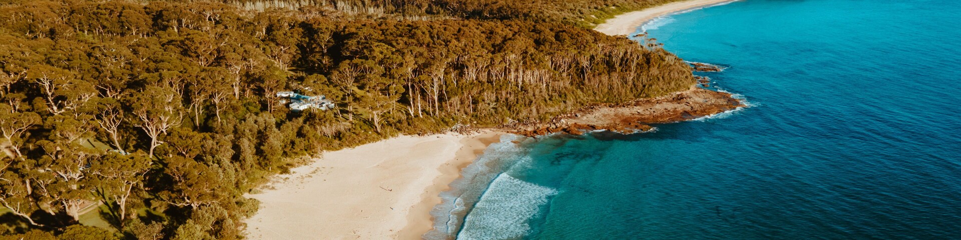 Paisagem costeira com uma praia arenosa, águas azuis cristalinas e colinas florestadas ao fundo.