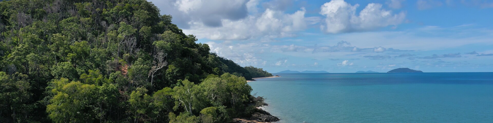 Paisagem costeira com uma praia arenosa, uma colina arborizada e um mar calmo sob um céu parcialmente nublado.