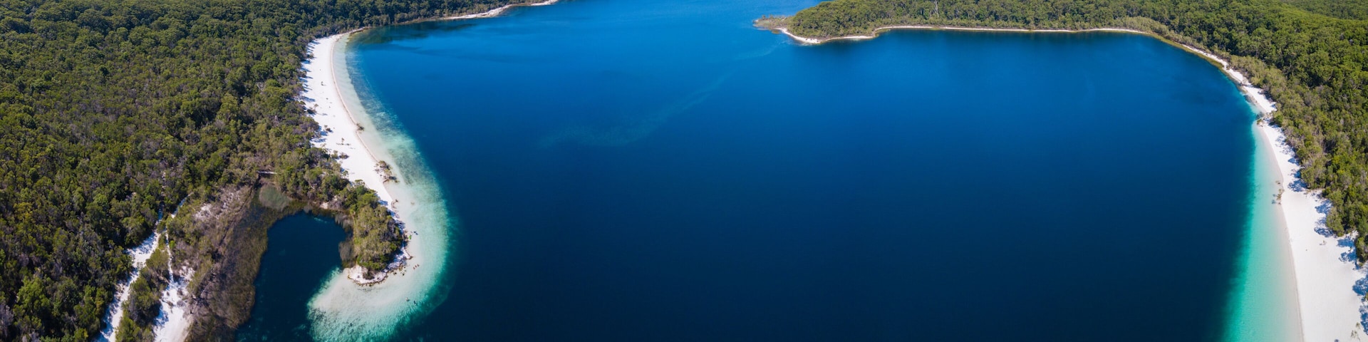 Um lago azul e translúcido, rodeado por uma floresta densa e praias de areia branca.