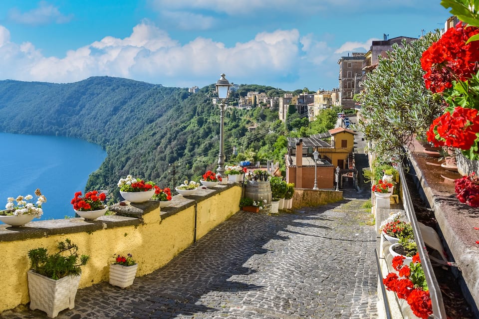 Ruas de pedra com vasos de flores e vista para um lago e colinas.
