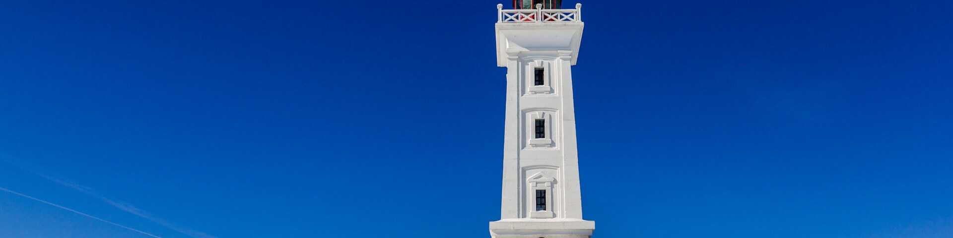 Um farol branco com topo vermelho, rodeado por uma paisagem coberta de neve sob um céu azul límpido.