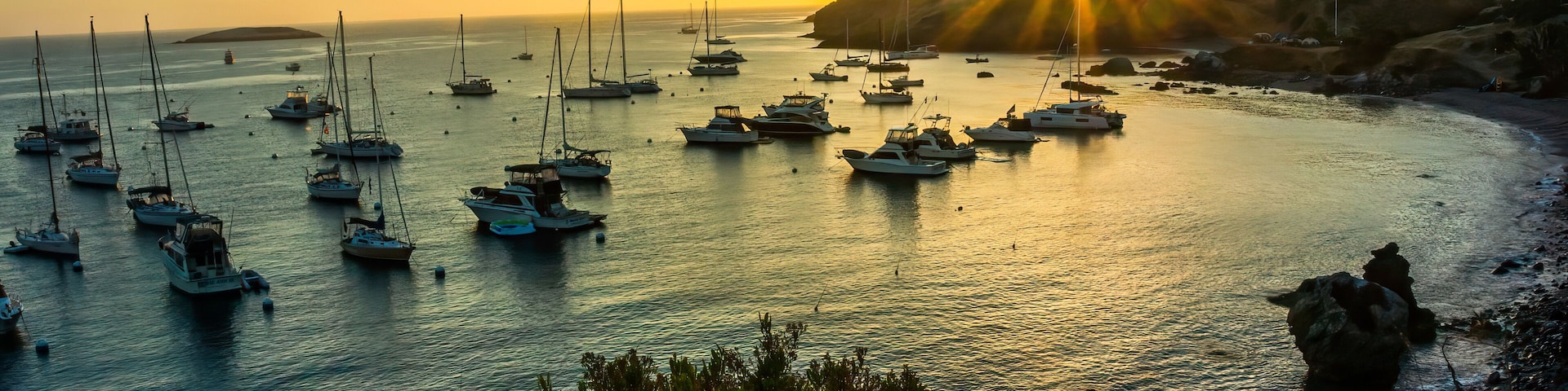 Cena costeira com barcos ancorados em águas calmas, um litoral rochoso e o sol se pondo ao fundo.