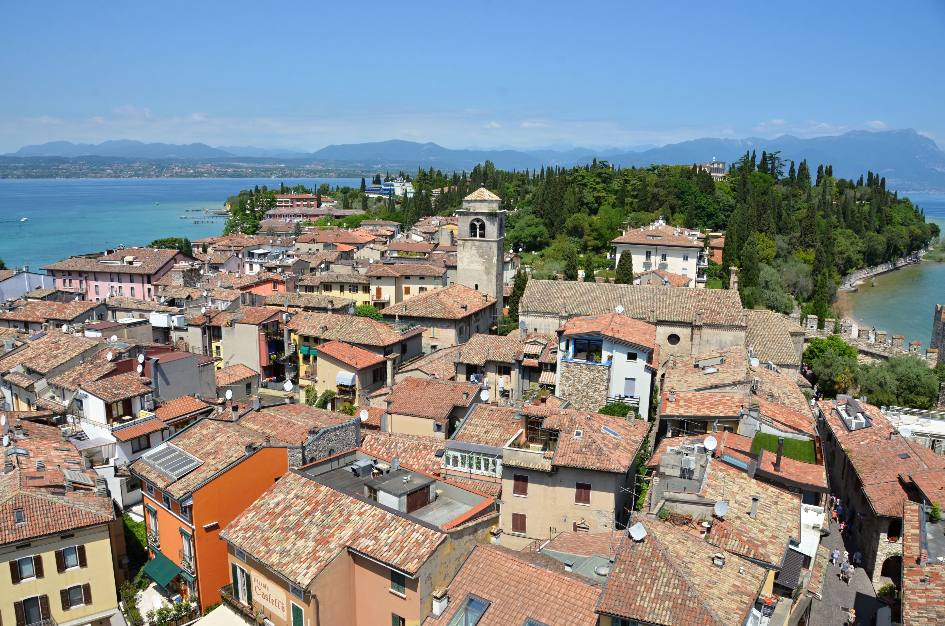Uma vila costeira com edifícios de telhados em terracota, uma torre de igreja imponente e uma vista clara do mar e montanhas ao longe.