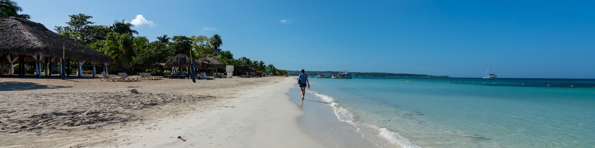 Uma praia com águas turquesa cristalinas, uma pessoa caminhando pela orla e estruturas com telhados de palha à esquerda.
