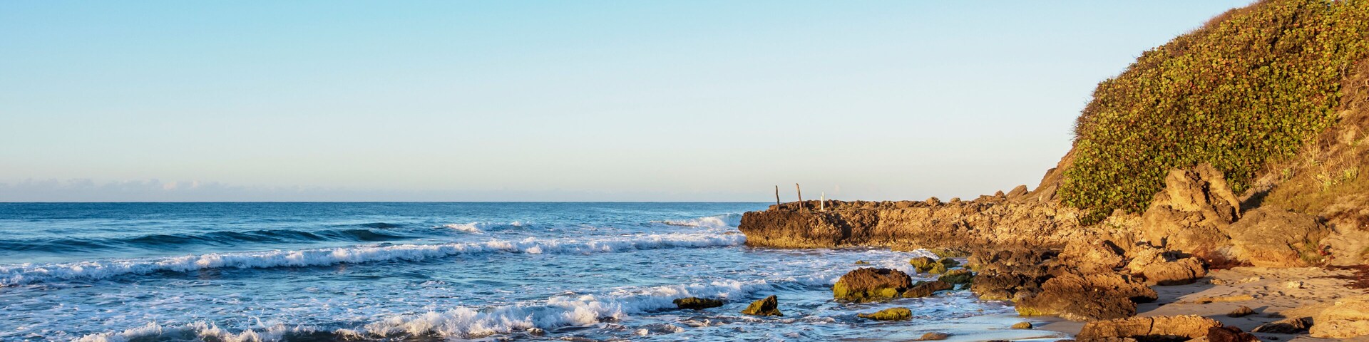 Litoral rochoso com ondas, uma praia de areia e uma colina com vegetação.