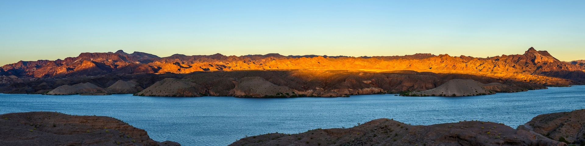 Paisagem montanhosa com um corpo d'água em primeiro plano e um pôr do sol projetando tons quentes nas colinas.