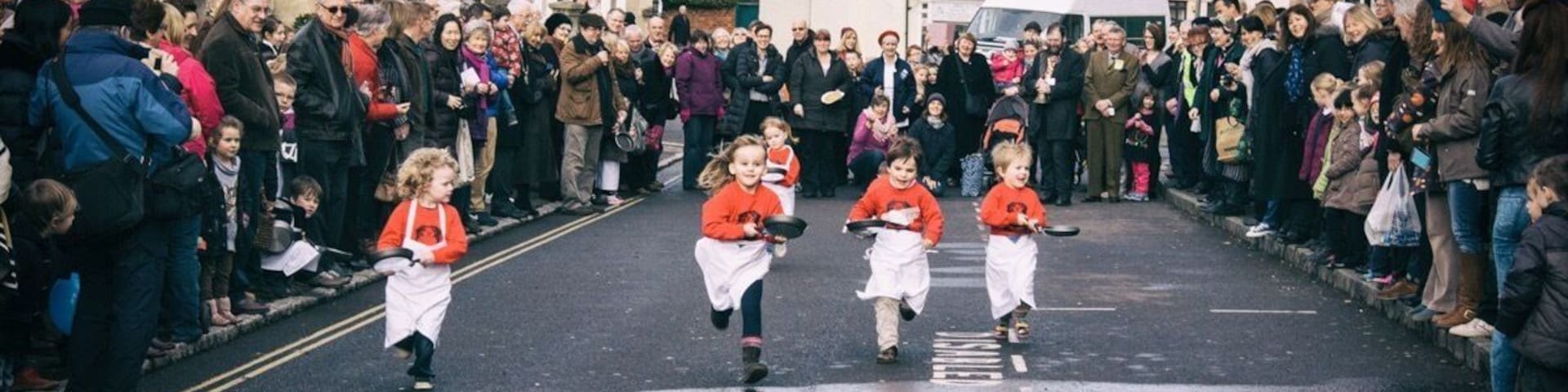 Um desfile de rua com crianças em uniformes laranja e branco correndo por uma estrada à beira de espectadores.